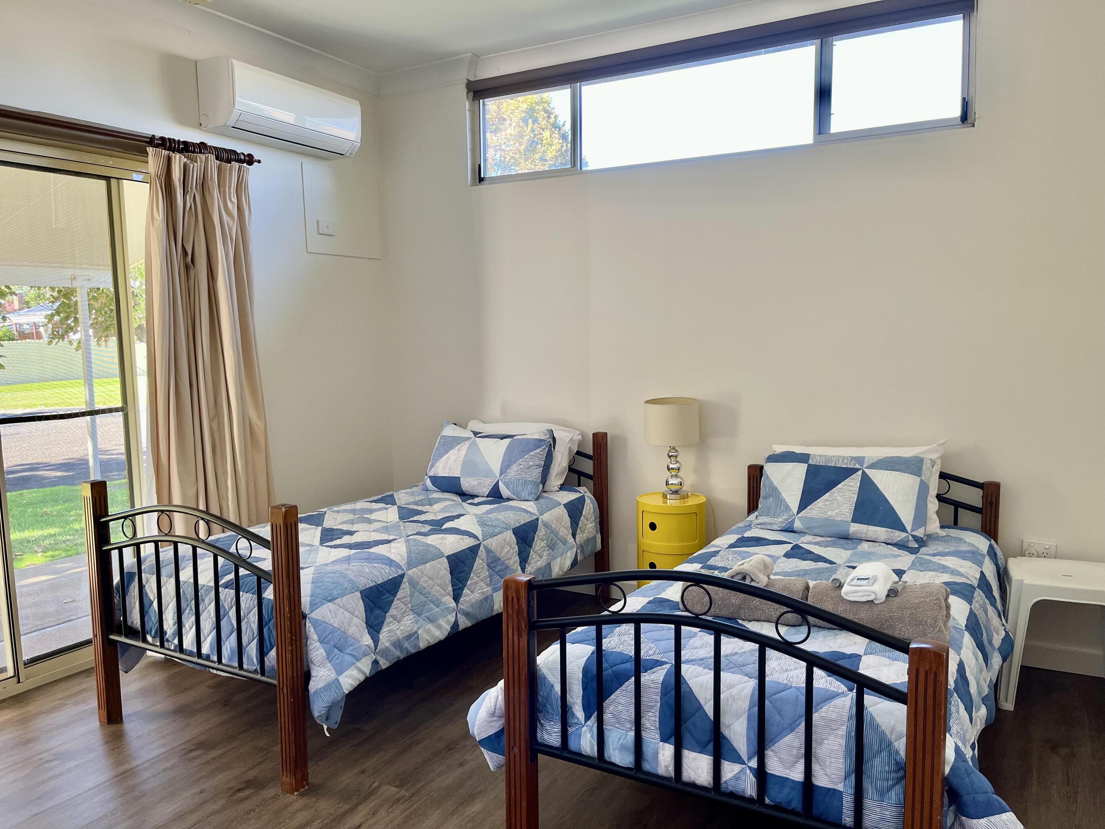 Quarto com duas camas, cada uma com colcha azul e branca, uma mesa de cabeceira amarela com abajur, e uma janela com vista para o verde.
