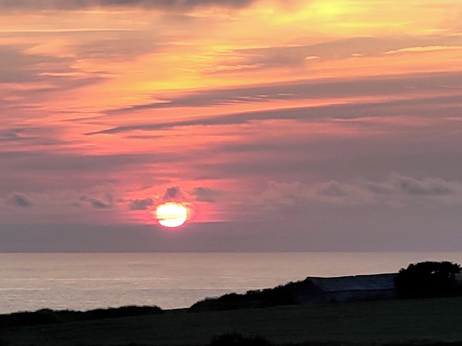 Um pôr do sol sobre uma paisagem costeira, com a silhueta de um edifício em primeiro plano.