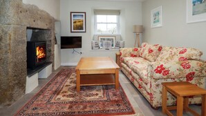 Uma sala de estar acolhedora com lareira de pedra, sofá estampado com flores, mesa de centro de madeira e televisão na parede.