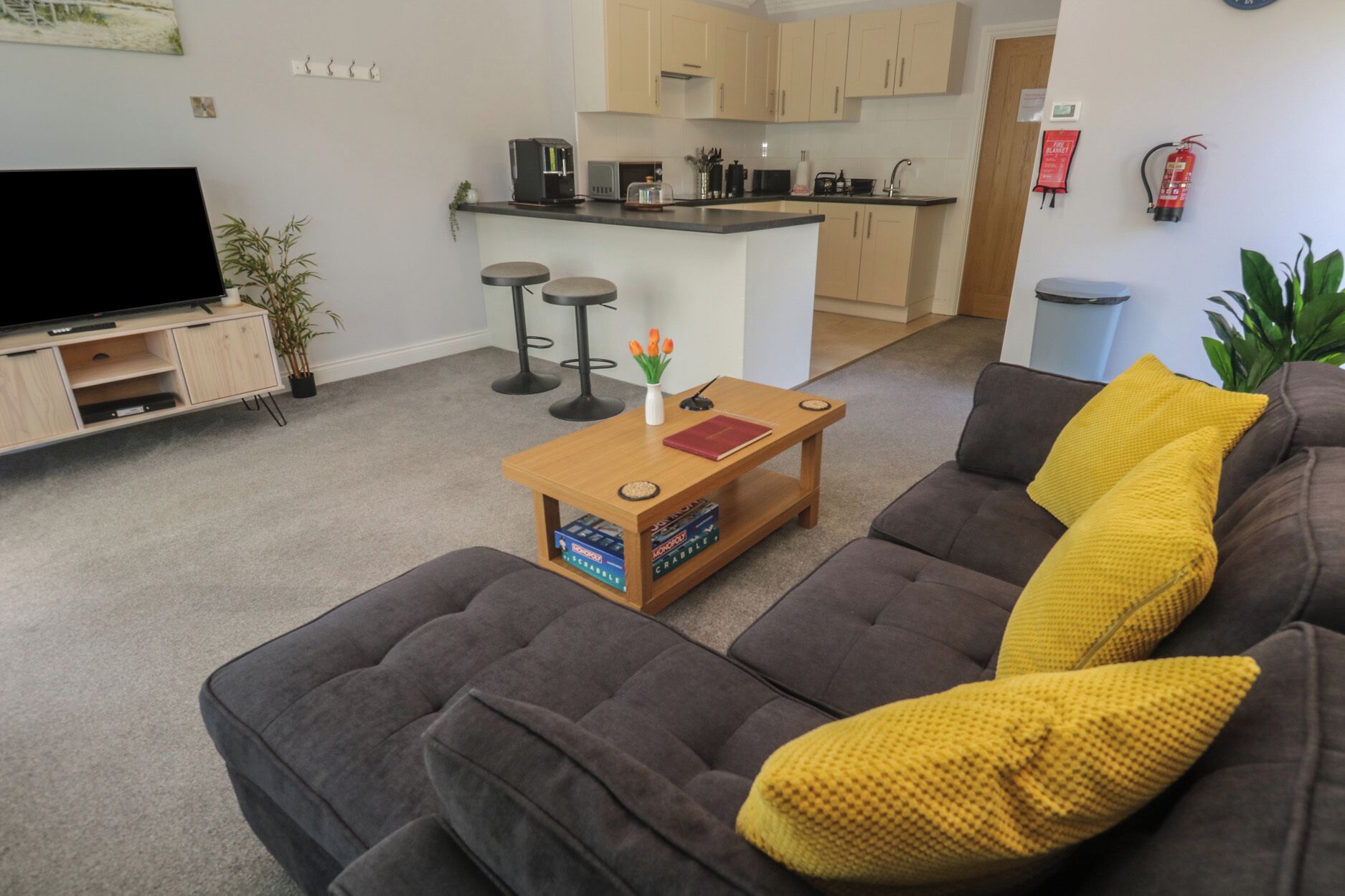 Uma sala de estar moderna com um sofá cinzento, uma mesa de centro de madeira e uma televisão em um suporte de madeira. Há uma área de cozinha com bancada preta e armários.