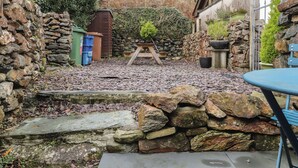 Um jardim com paredes de pedra, uma mesa e bancos de madeira, rodeado por plantas em vasos e uma estrutura de madeira.