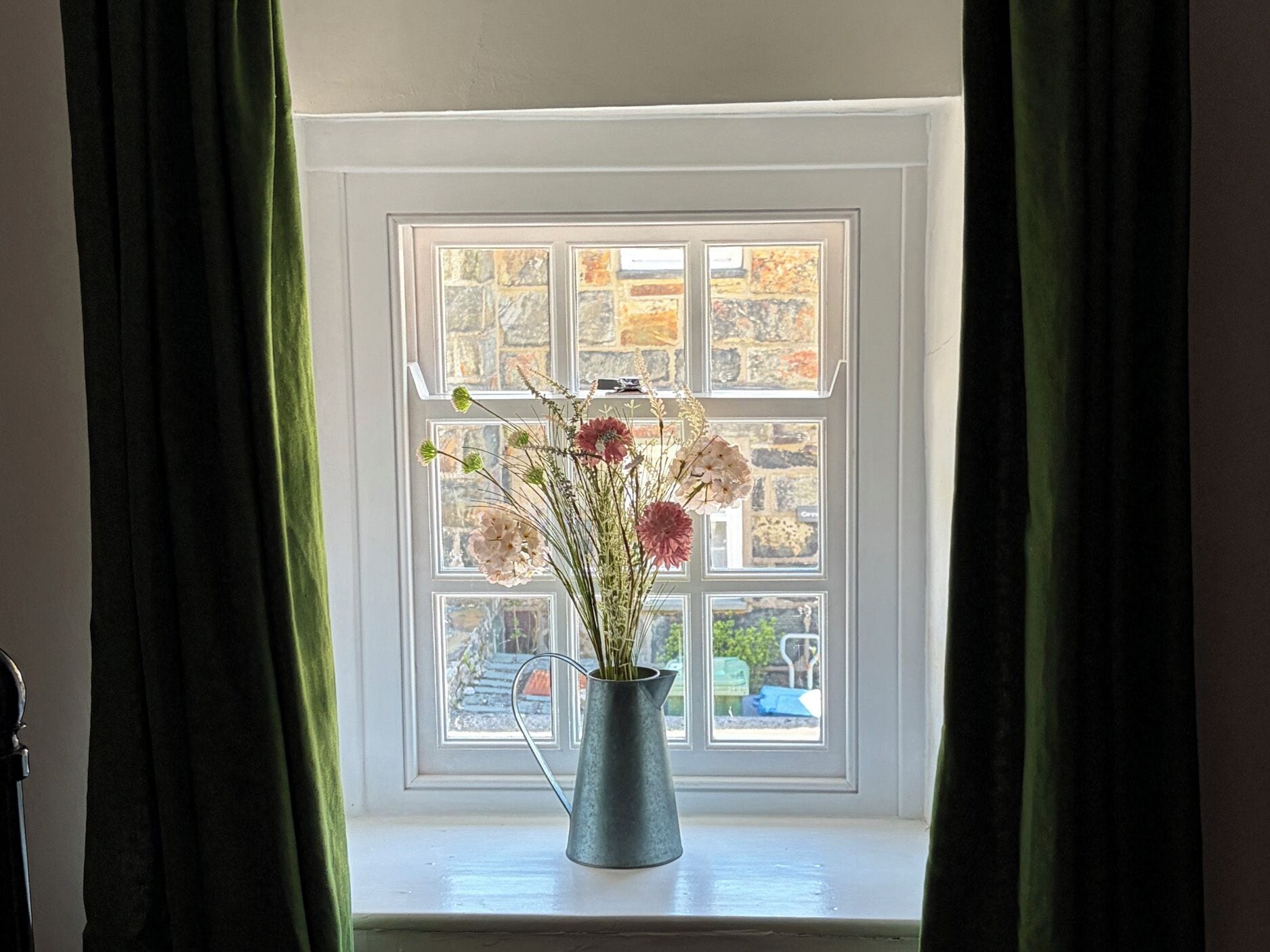 Uma janela com vista para uma parede de pedra, um vaso de metal com flores e cortinas verdes.