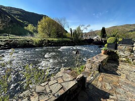 Uma ponte de pedra sobre um rio corrente, com vista para uma colina e árvores ao fundo.