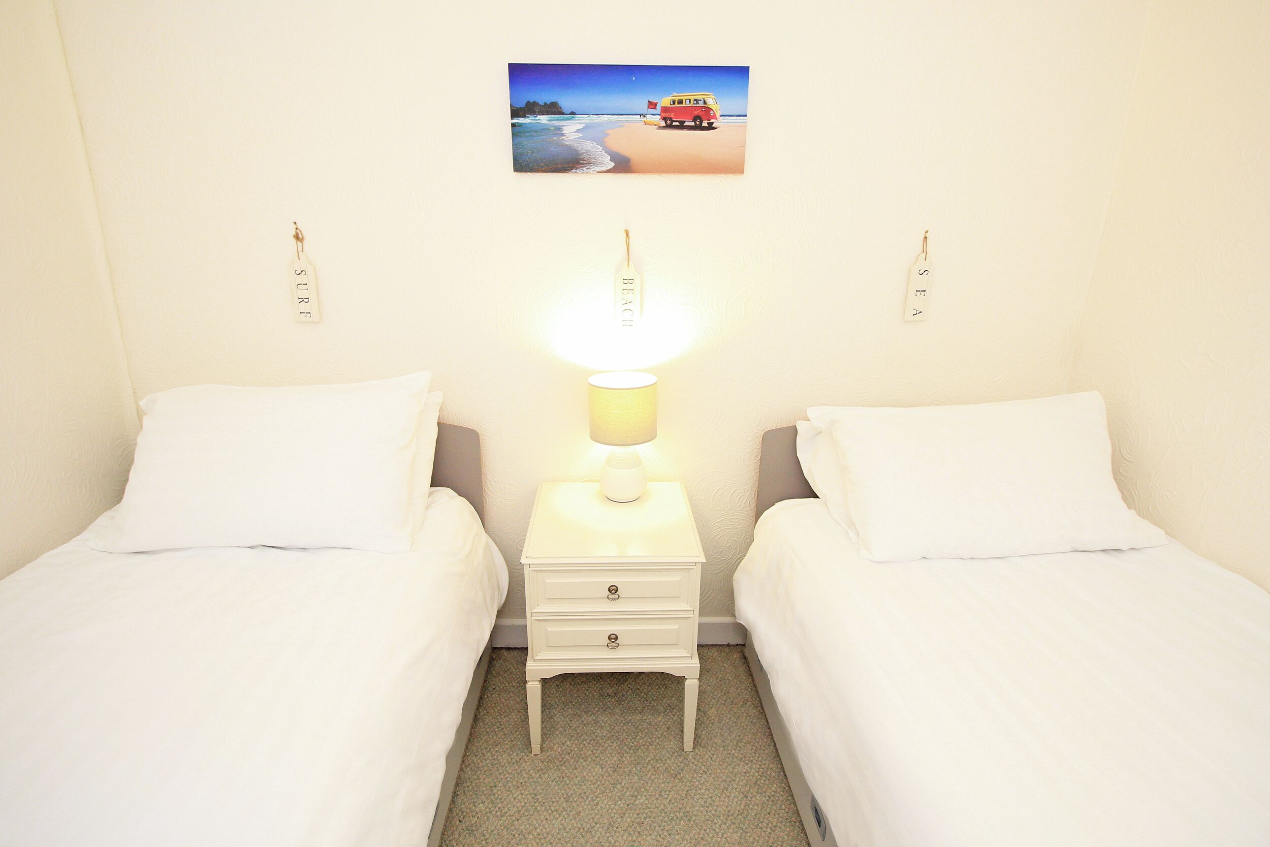 Quarto de hotel com duas camas de solteiro, uma mesa de cabeceira com abajur e um quadro com tema de praia na parede.