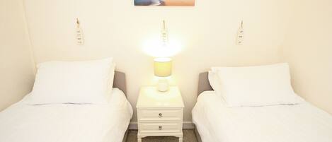 Quarto de hotel com duas camas de solteiro, uma mesa de cabeceira com abajur e um quadro com tema de praia na parede.