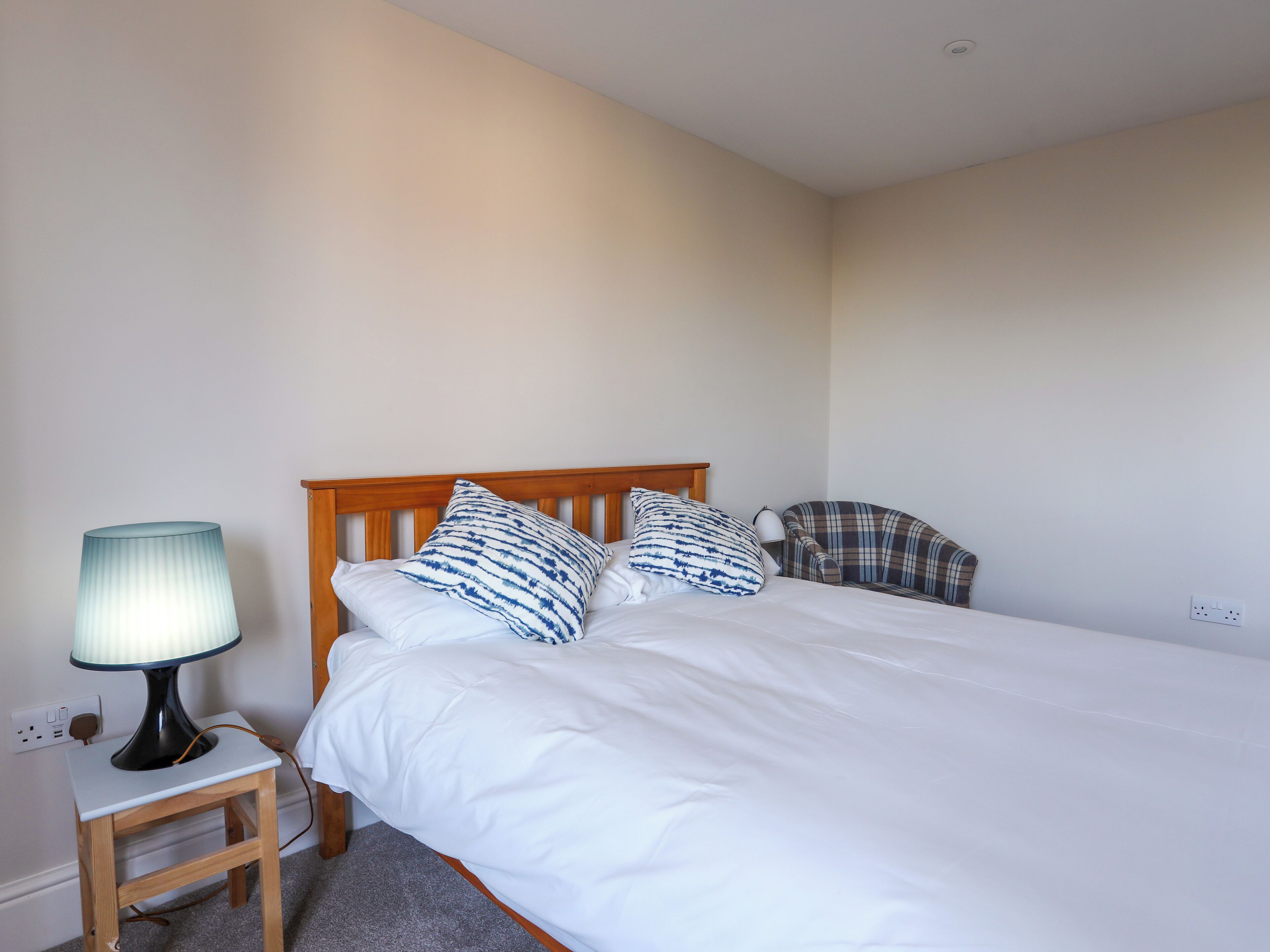 Cama bem arrumada com roupa de cama branca e almofadas listradas, uma poltrona xadrez, um pequeno criado-mudo de madeira com uma luminária e uma tomada na parede.
