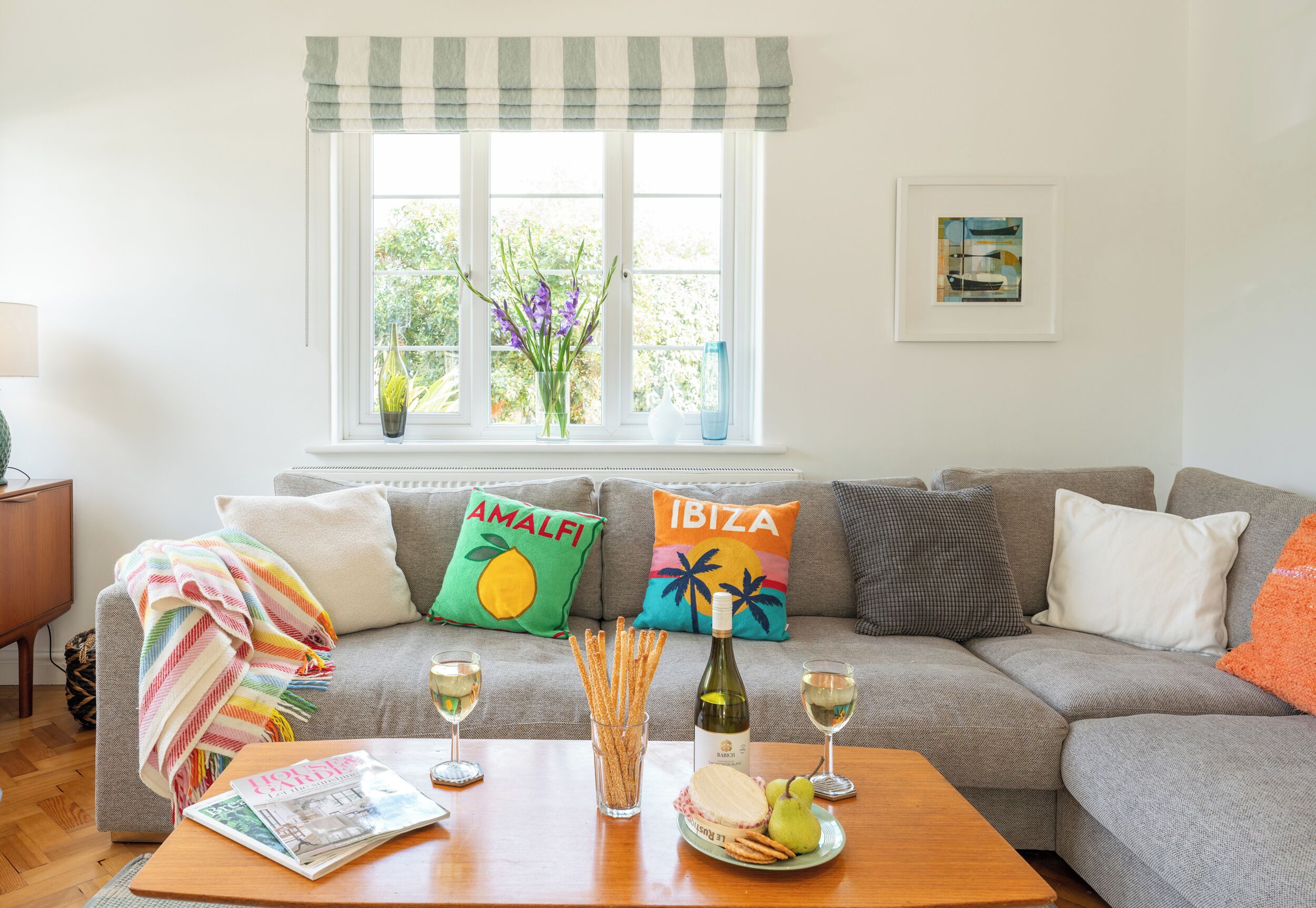 Uma sala de estar aconchegante com um sofá, almofadas coloridas, uma janela com vista, uma mesa com bebidas, petiscos e uma revista.