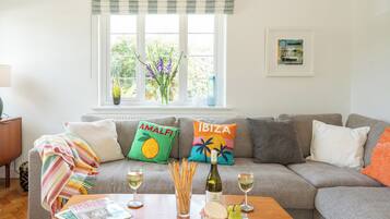 Uma sala de estar aconchegante com um sofá, almofadas coloridas, uma janela com vista, uma mesa com bebidas, petiscos e uma revista.