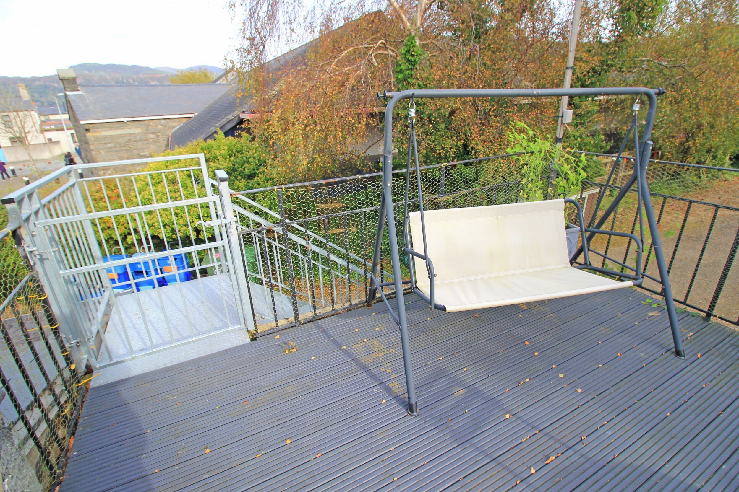 Um balanço de metal sobre um deck de madeira, com uma cerca de metal e um recipiente azul ao fundo.