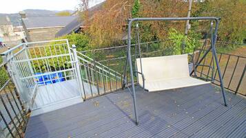 Um balanço de metal sobre um deck de madeira, com uma cerca de metal e um recipiente azul ao fundo.