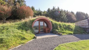 Estrutura circular de tijolos com porta de vidro, cercada por área gramada e árvores.
