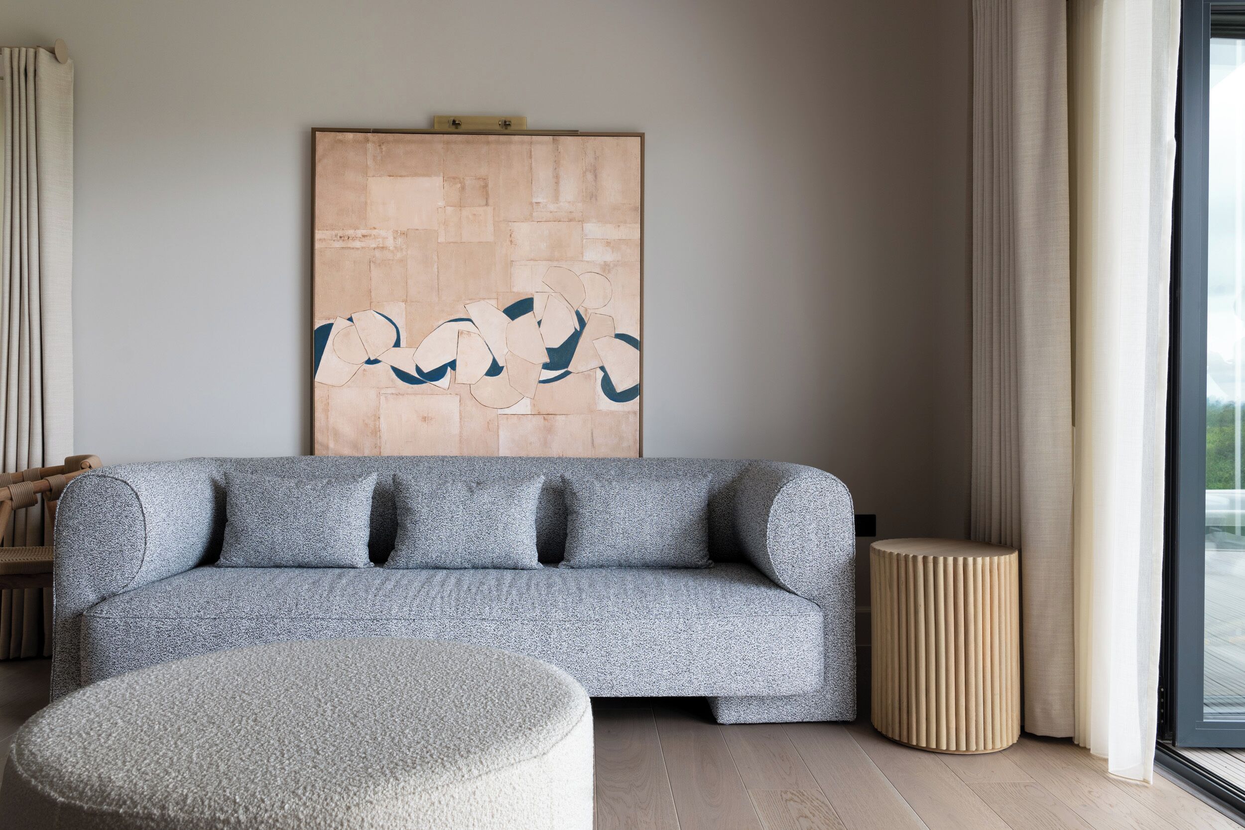 Uma sala de estar moderna com um sofá cinza, um banquinho redondo, uma mesa de centro de madeira e uma grande pintura abstrata na parede.