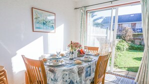 Uma área de refeições com uma mesa posta para quatro pessoas, cadeiras de madeira, toalha de mesa com motivos náuticos, um vaso com flores e vista para um jardim por uma porta deslizante.