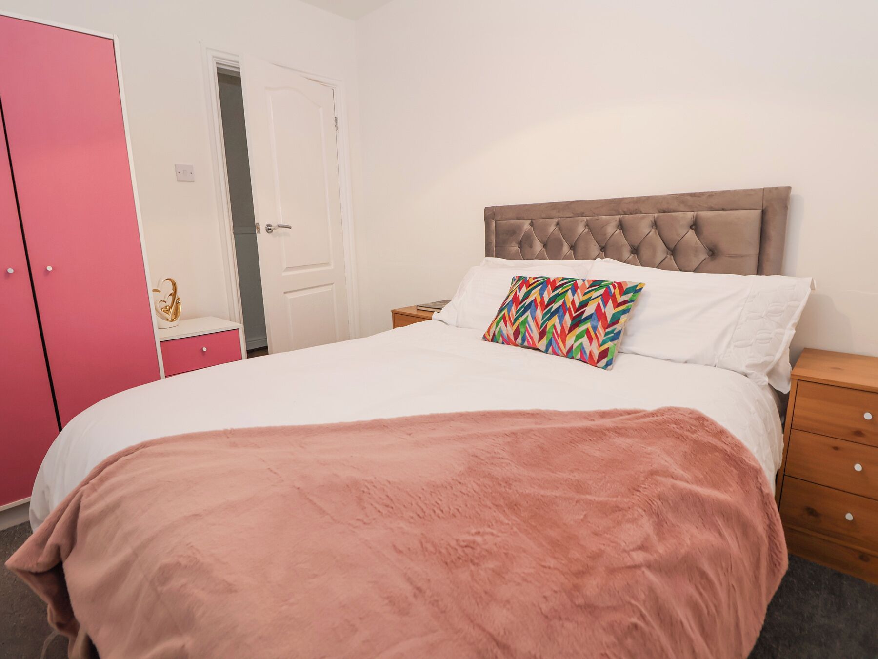 Quarto com uma cama grande, um guarda-roupa rosa, uma mesa de cabeceira de madeira e uma porta.