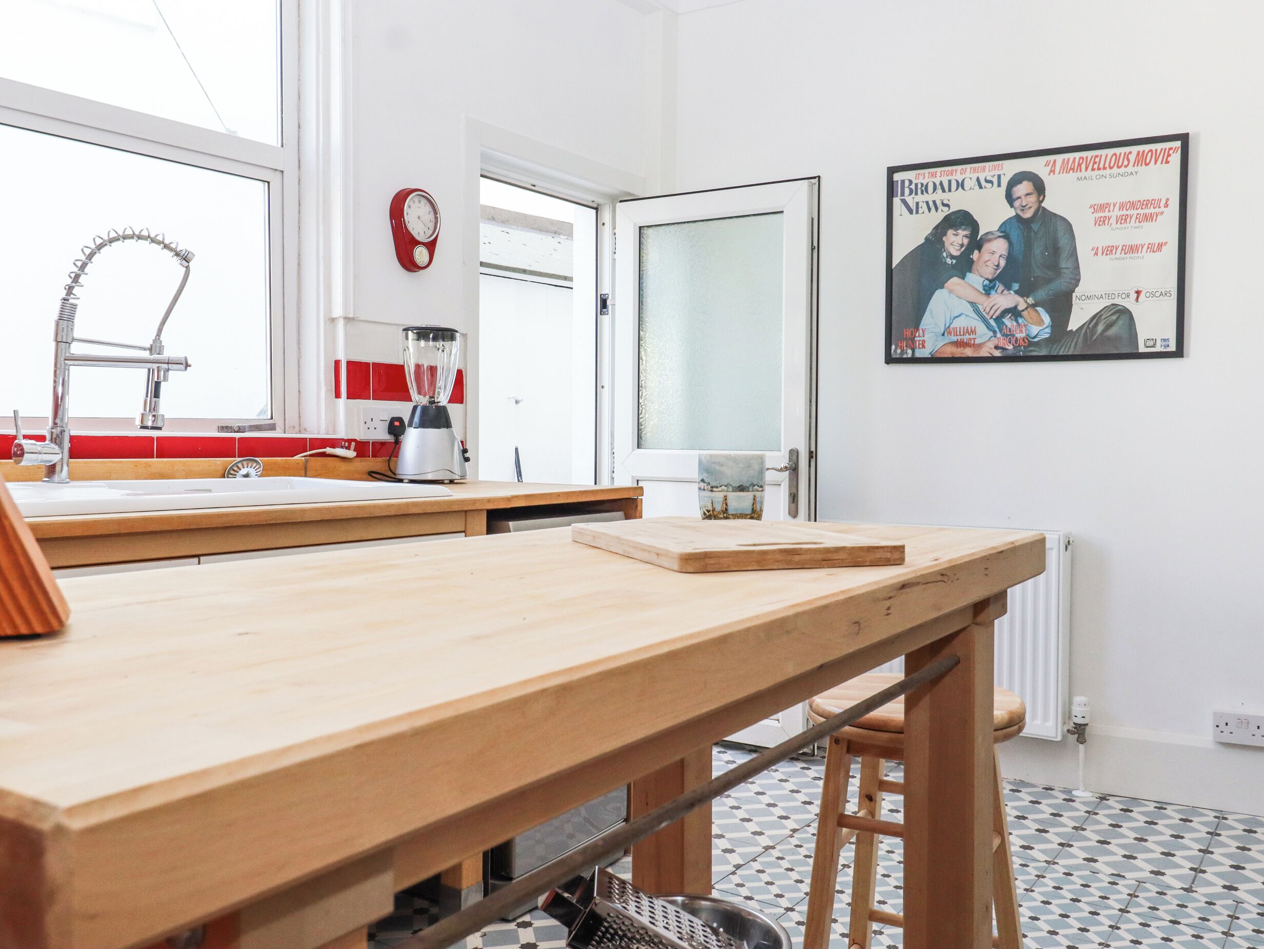 Uma cozinha com uma mesa de madeira, um liquidificador, uma chaleira e um relógio na parede.