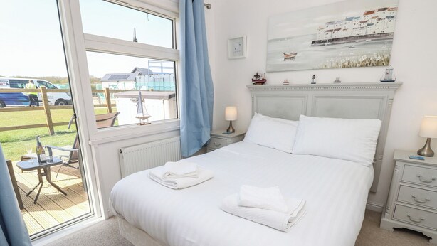 Quarto de hotel com uma cama grande, uma janela com vista para o exterior, uma mesa pequena com uma garrafa e copos, e um quadro na parede.