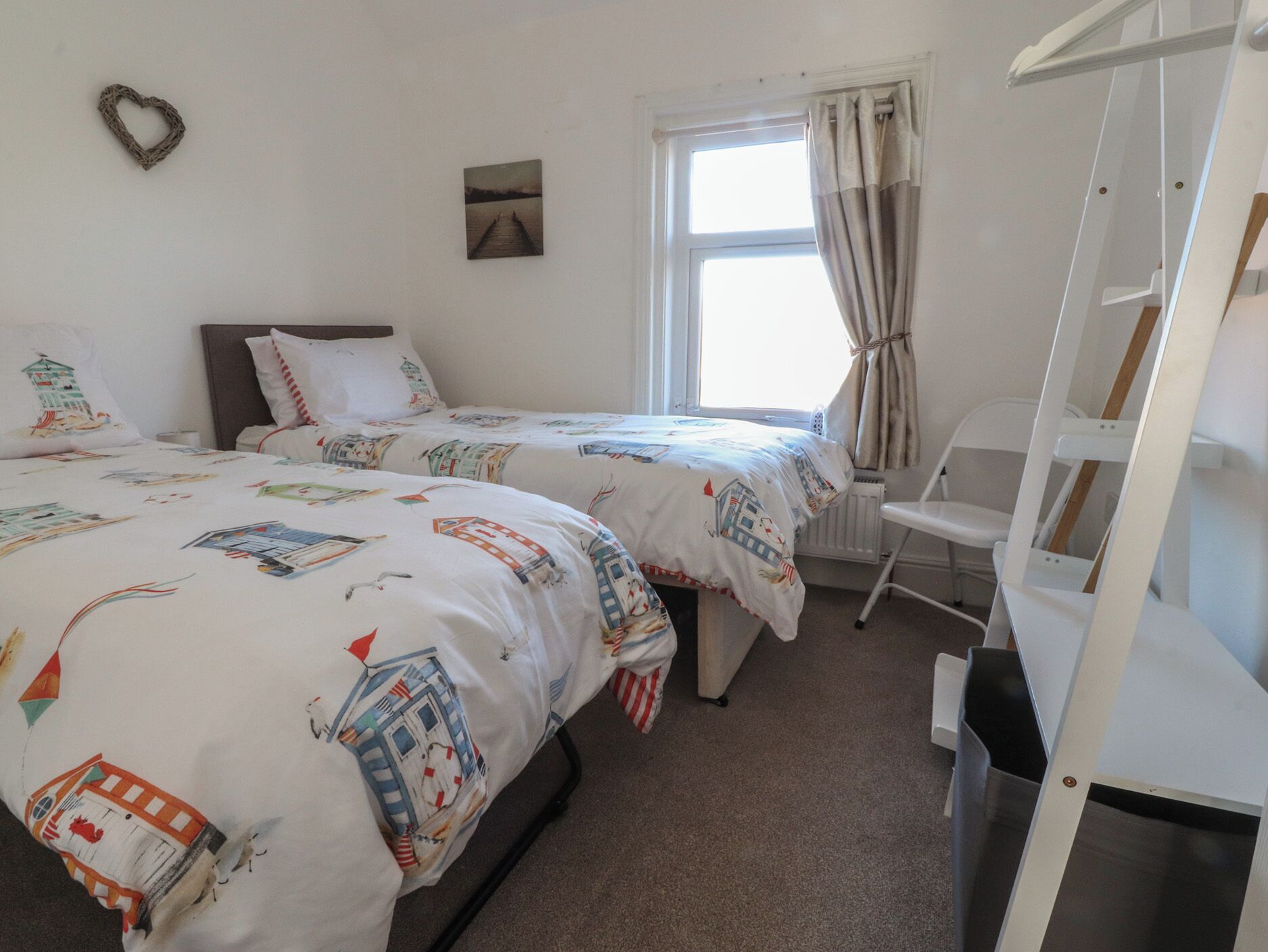 Quarto com duas camas, janela com cortinas, radiador e uma cadeira branca.