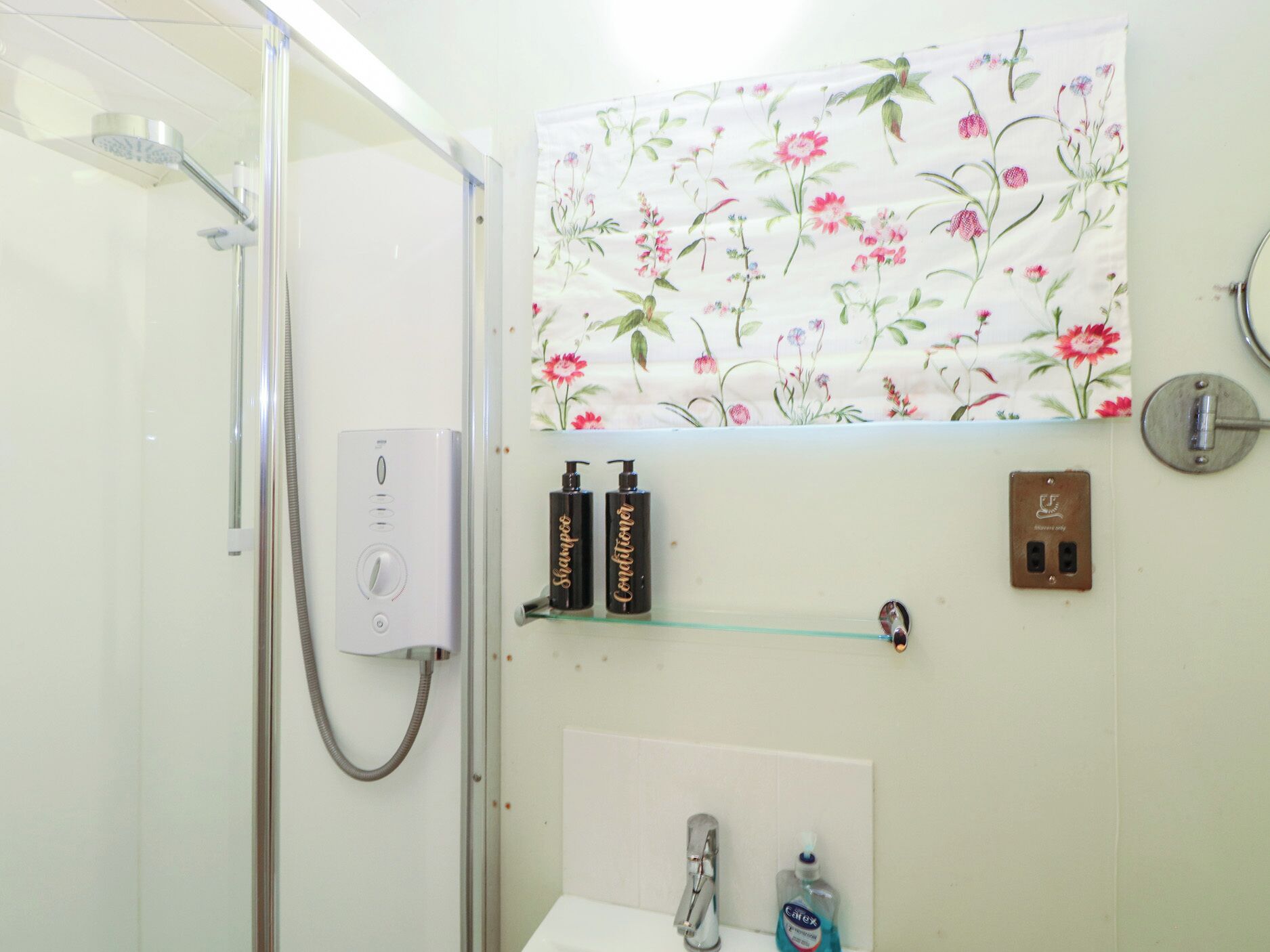 Chuveiro com painel de controle, espelho com papel de parede floral e produtos de higiene em uma prateleira.