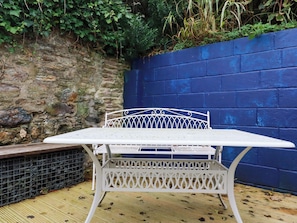 Mesa de metal branca com encosto decorativo, sobre um deck de madeira com parede de pedra e parede azul ao fundo.