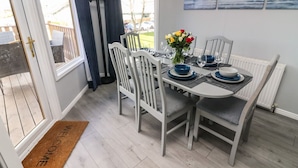 Uma sala de jantar com uma mesa posta para quatro pessoas, cadeiras, um vaso com flores e vista para o exterior através de uma janela.