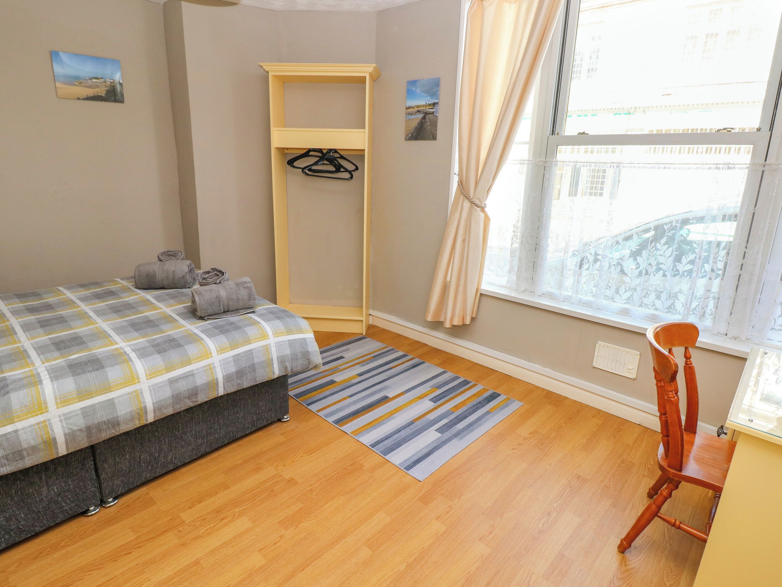 Quarto com cama, piso de madeira, cadeira, janela com cortinas e prateleira fixada na parede com suporte para casacos.