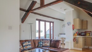 Uma sala de estar ampla com um sofá de couro marrom, uma mesa de centro de vidro e piso de madeira. Há uma área de cozinha com armários de madeira e um extintor de incêndio vermelho.