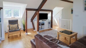 Um amplo loft com vigas de madeira aparentes, uma área de estar aconchegante com sofá e poltrona de couro, uma televisão e uma estante de livros.