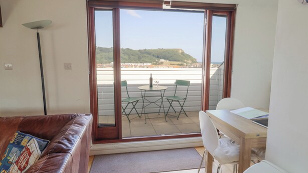 Quarto com porta de vidro deslizante que dá para uma varanda com mesa e cadeiras, uma escrivaninha de madeira, um sofá de couro marrom e um tapete pequeno.
