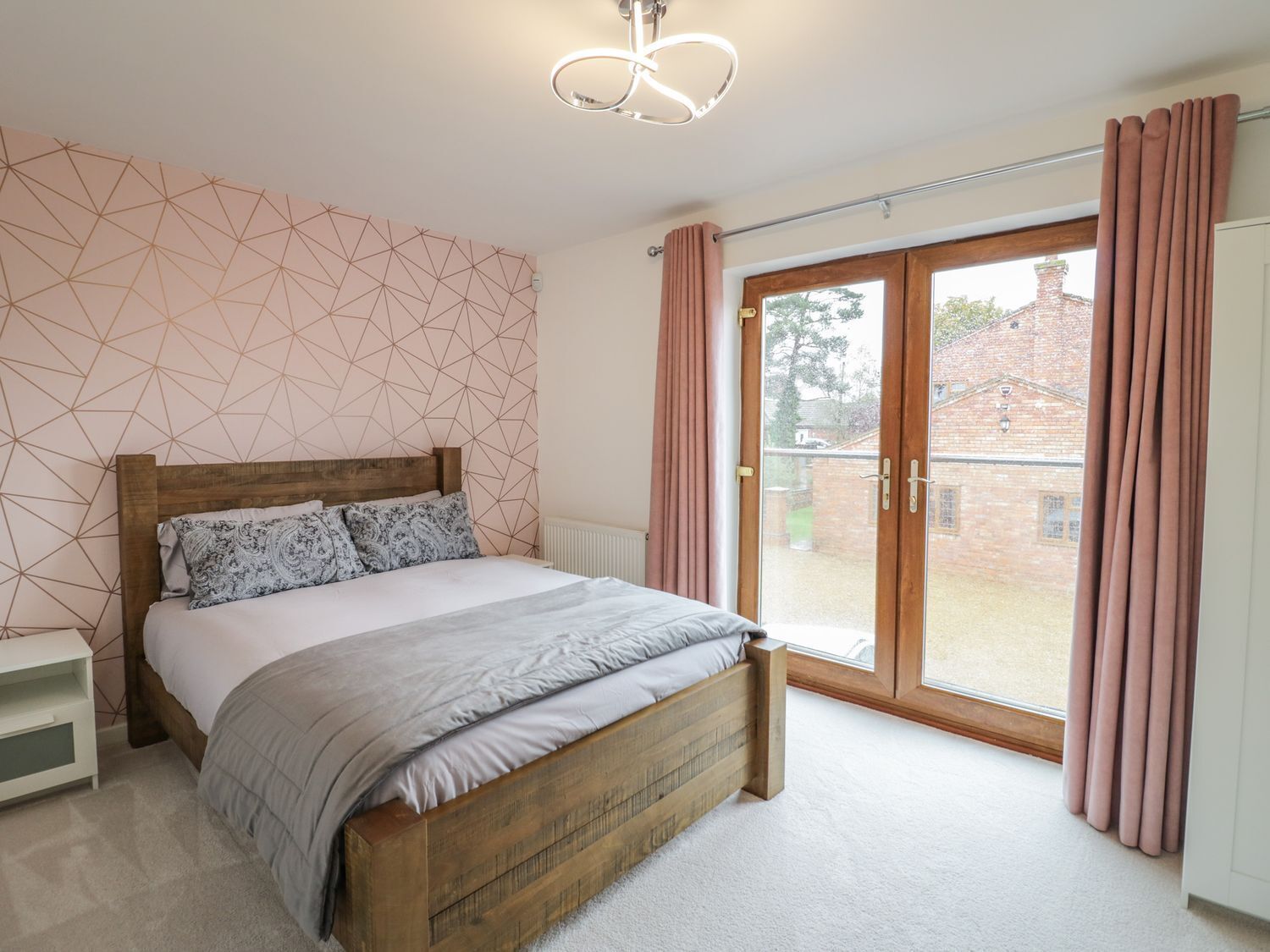 Quarto com cama de madeira, almofadas estampadas, design geométrico na parede, mesa de cabeceira branca e vista para área externa por uma porta de vidro deslizante.