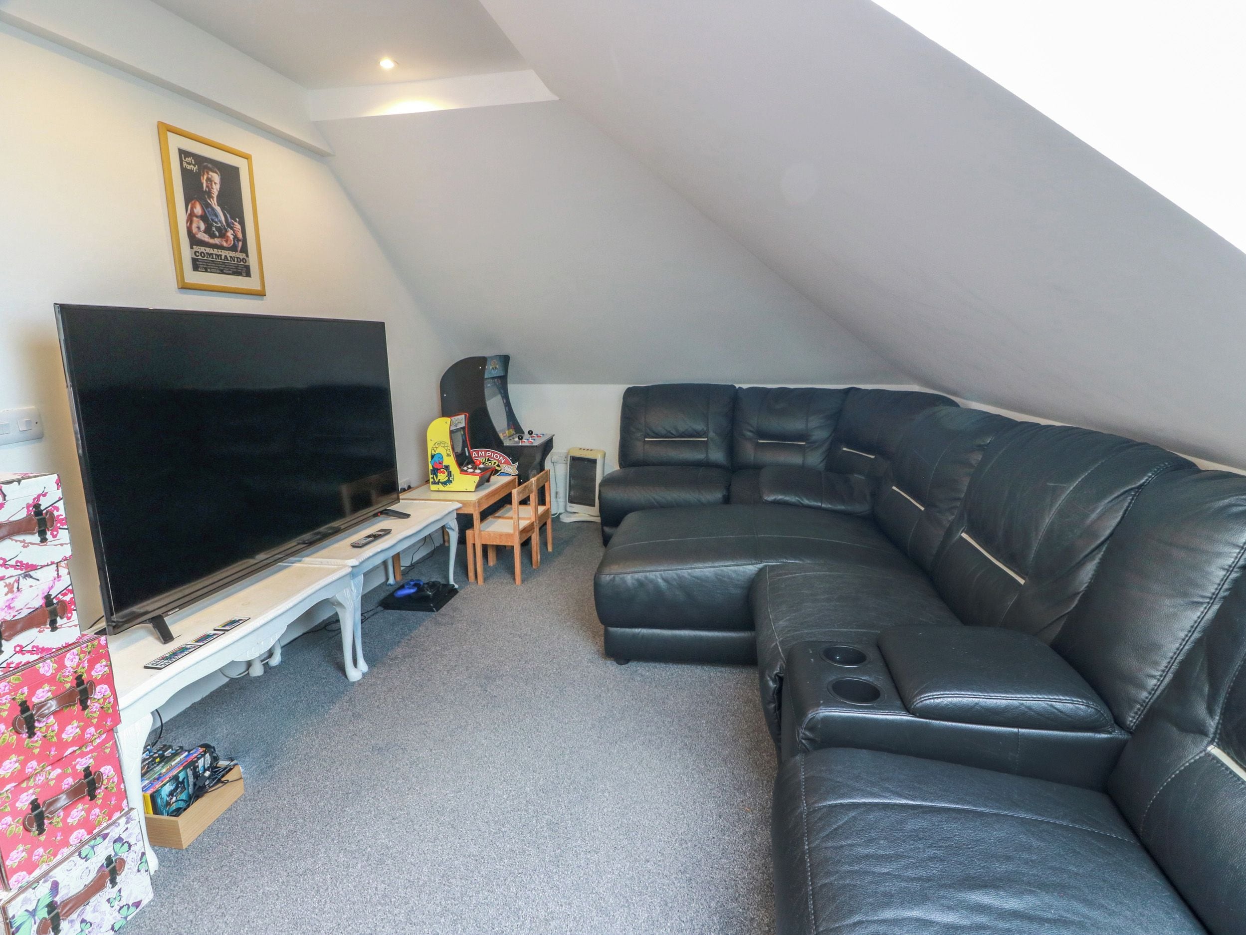 Uma sala de estar com uma televisão de tela plana grande, um console de jogos, um sofá de couro preto e uma mesa pequena.