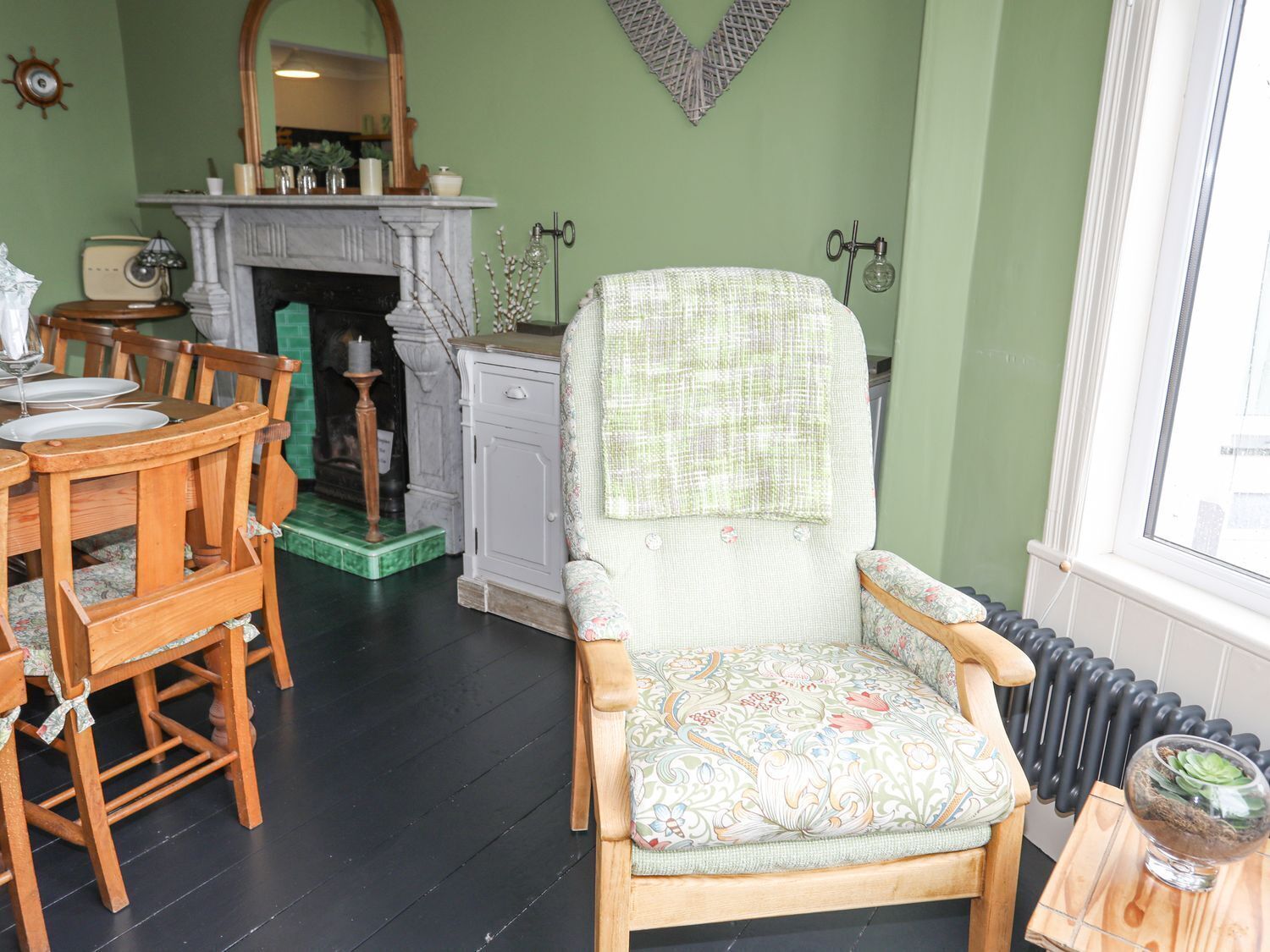 Quarto com parede verde, lareira, mesa de jantar com cadeiras e poltrona estofada floral.