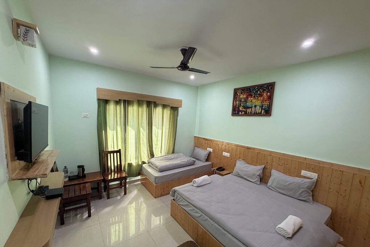 Quarto de hotel com duas camas, uma escrivaninha com computador, uma cadeira, televisão e ventoinha de teto.