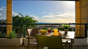 Varanda com mesa posta para o café da manhã, com vista para a costa, palmeiras e céu claro.
