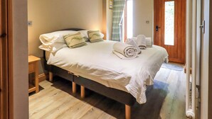 Cama bem arrumada com lençóis brancos e toalhas dobradas, piso de madeira e porta de madeira em um quarto com paredes bege.