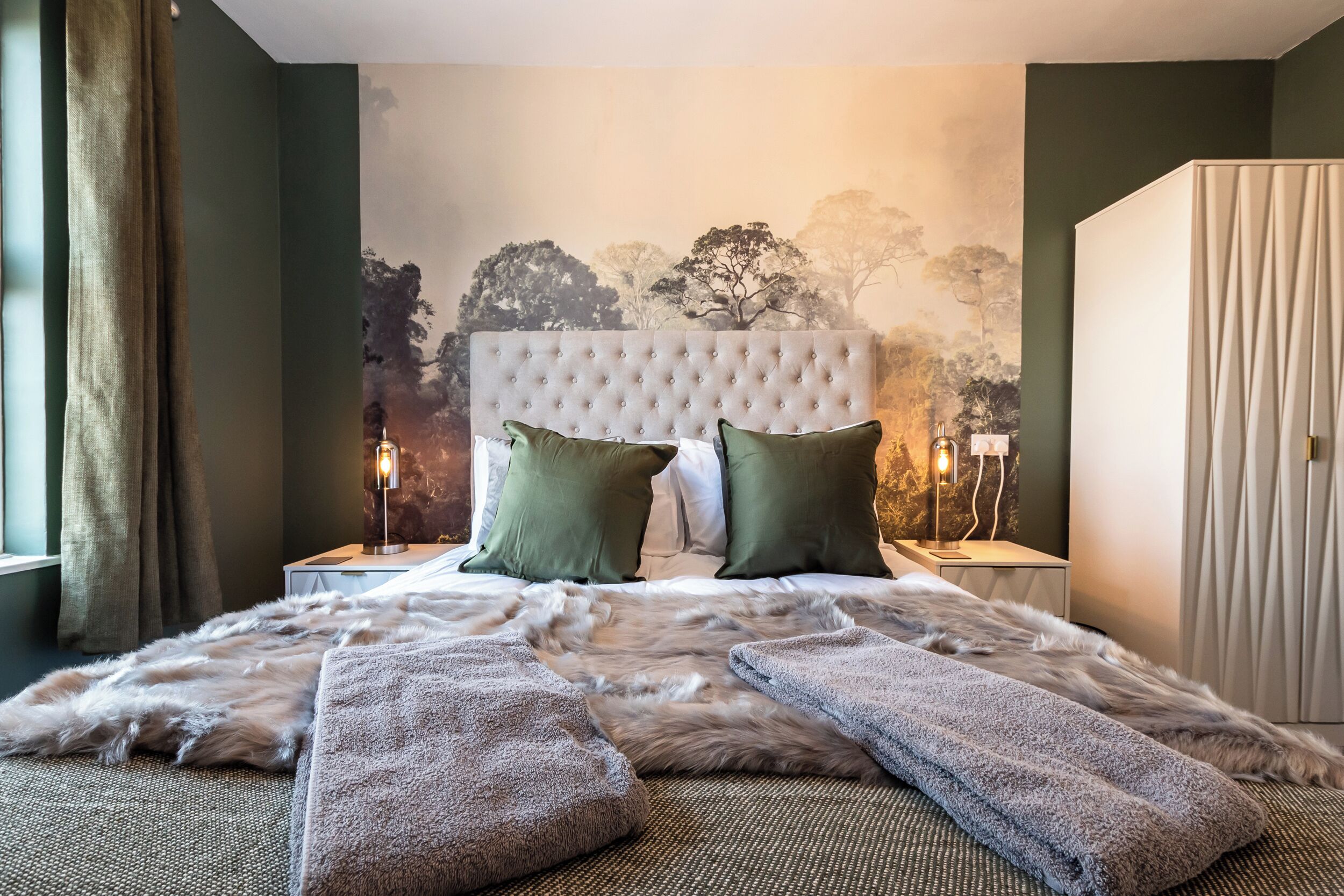 Quarto com cabeceira estofada, duas almofadas verdes, manta de pele e mesa de cabeceira com abajur.