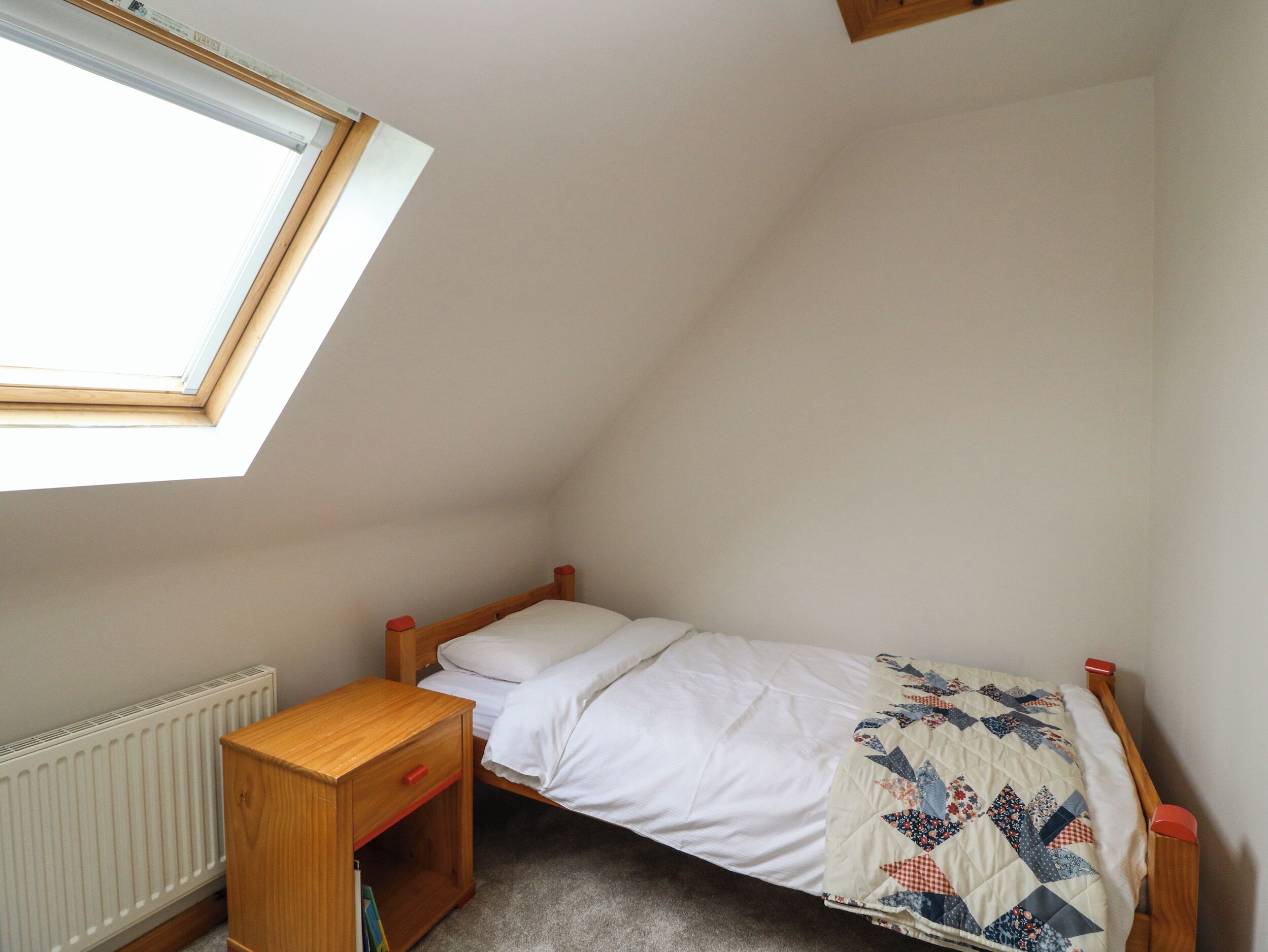 Quarto pequeno com uma cama de solteiro, teto inclinado, mesa de cabeceira de madeira, radiador e claraboia.