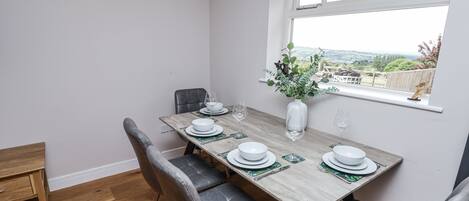 Uma sala de jantar com uma mesa de madeira posta para quatro pessoas, cadeiras estofadas cinzentas e vista para o campo através de uma janela.