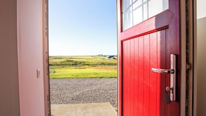 Uma porta vermelha dá acesso a um caminho de cascalho, com vista para uma paisagem verde e um edifício ao longe.