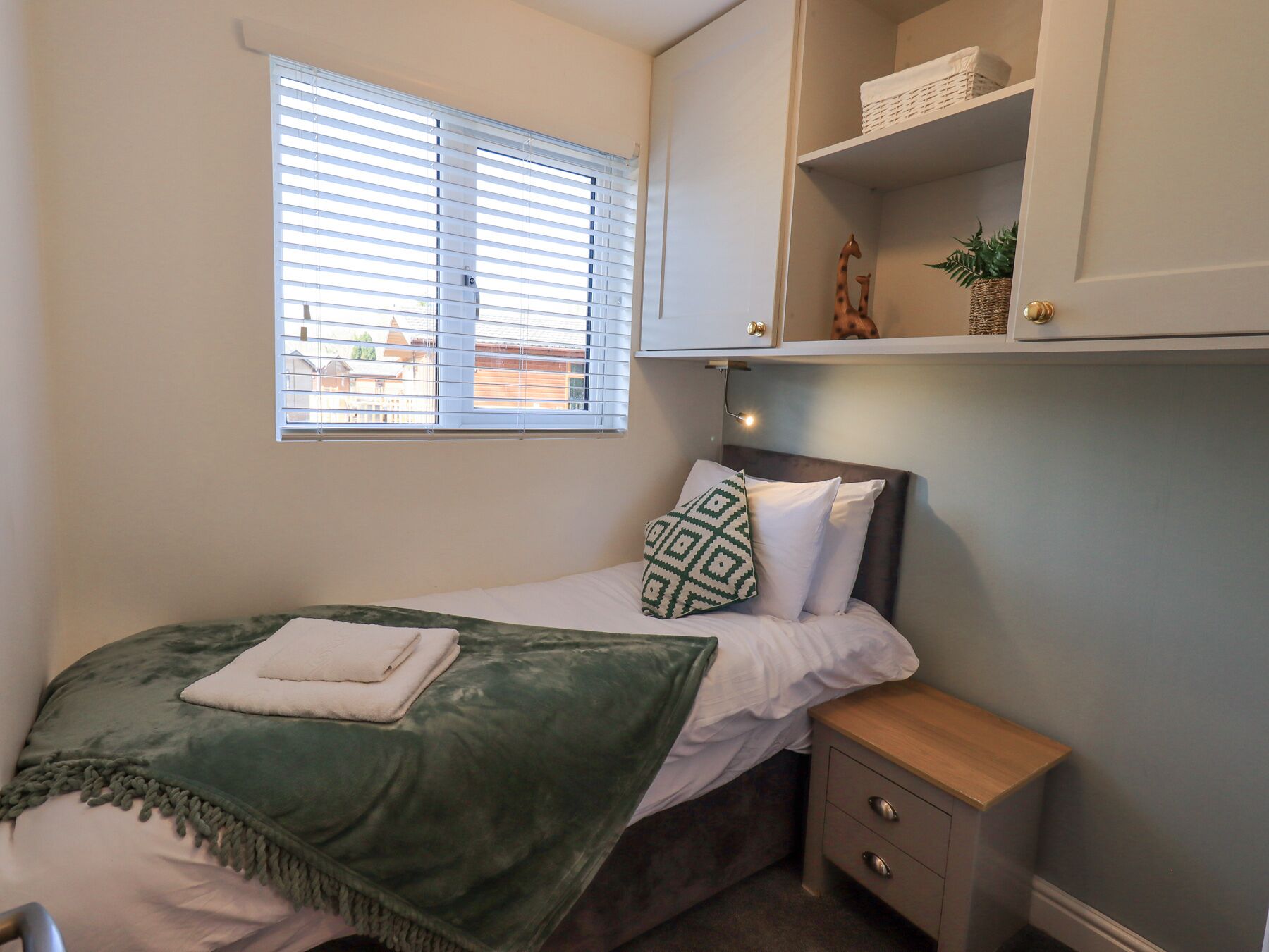 Quarto com cama, mesa de cabeceira, estante, janela com persianas e prateleira com objetos decorativos.