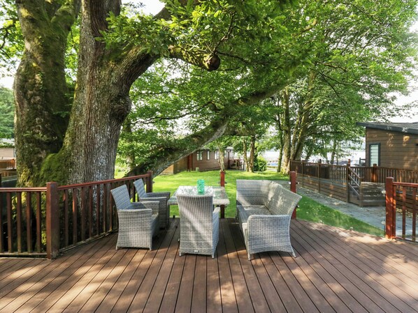 Um deck de madeira com móveis de vime, uma mesa de vidro e vista para um lago e edifícios ao fundo.