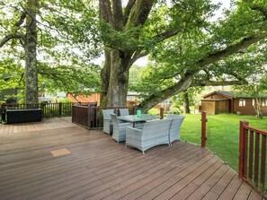 Um deck de madeira com móveis de vime e uma mesa, cercado por árvores altas e um gramado.