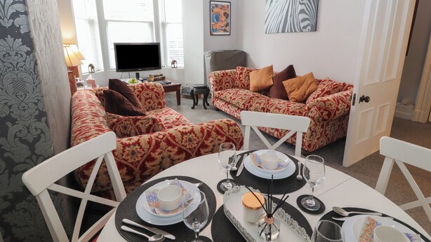 Uma sala de estar com um sofá, uma mesa de jantar posta para quatro pessoas e uma televisão ao fundo.