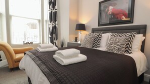 Cama bem arrumada com colcha escura, travesseiros brancos e toalhas dobradas. Há uma cadeira, um abajur e um quadro na parede.