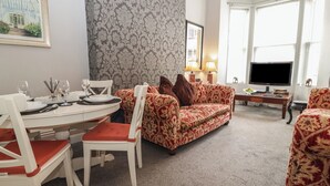 Uma sala de estar com uma mesa branca posta para quatro pessoas, um sofá estampado e uma televisão, além de um tapete padronizado.