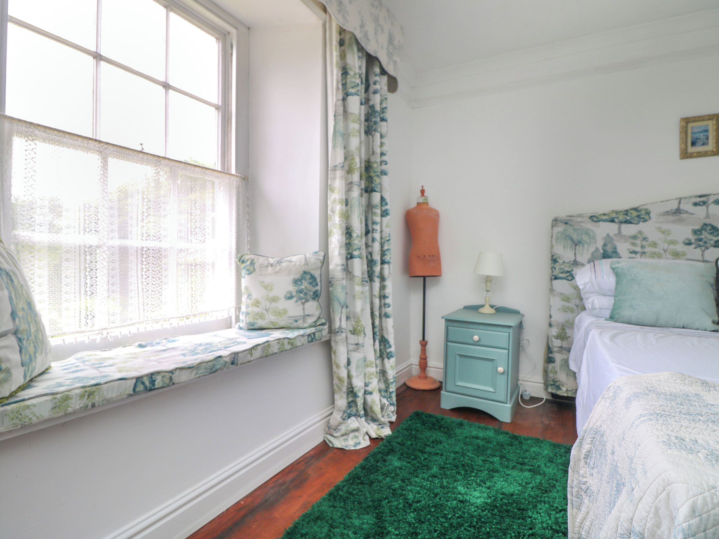 Quarto com um assento junto à janela, cama com roupa de cama florida, mesa de cabeceira com abajur e tapete verde.