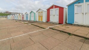Uma fileira de cabanas de praia coloridas, com portas brancas e numeradas 104, numa área pavimentada com uma colina gramada ao fundo.