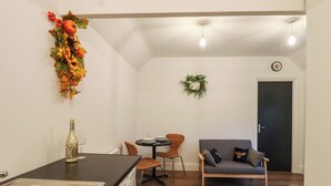 Uma cozinha compacta com pia e área de jantar com uma mesa redonda e duas cadeiras. Um sofá está posicionado ao fundo, e uma guirlanda decorativa pendurada na parede.