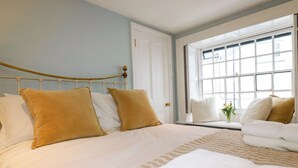 Cama bem arrumada com roupa de cama branca e bege, janela com vista para o exterior e porta branca.