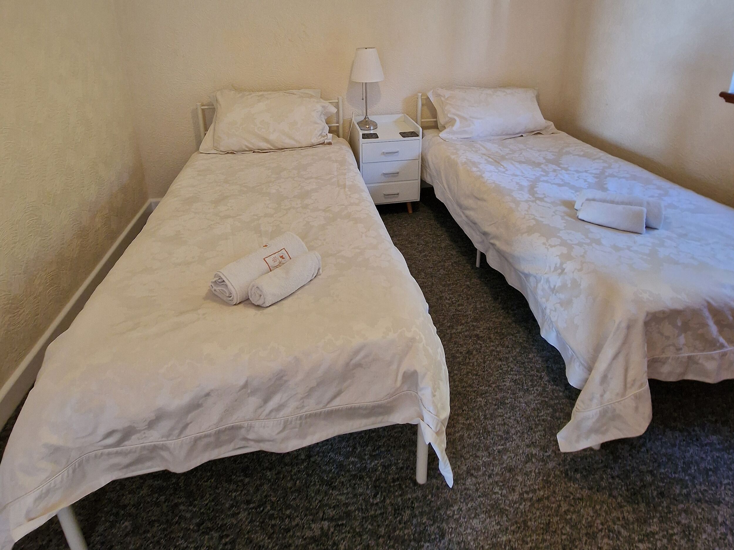 Quarto com duas camas de solteiro, cada uma com travesseiro e toalha enrolada. Há uma mesinha de cabeceira com abajur entre as camas.