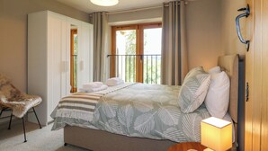 Quarto com cama, cadeira, mesa de cabeceira, abajur e vista para o exterior pela janela.